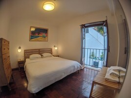 Quarto com cama de madeira, mesa de cabeceira, cômoda, pintura e varanda com plantas em vasos.