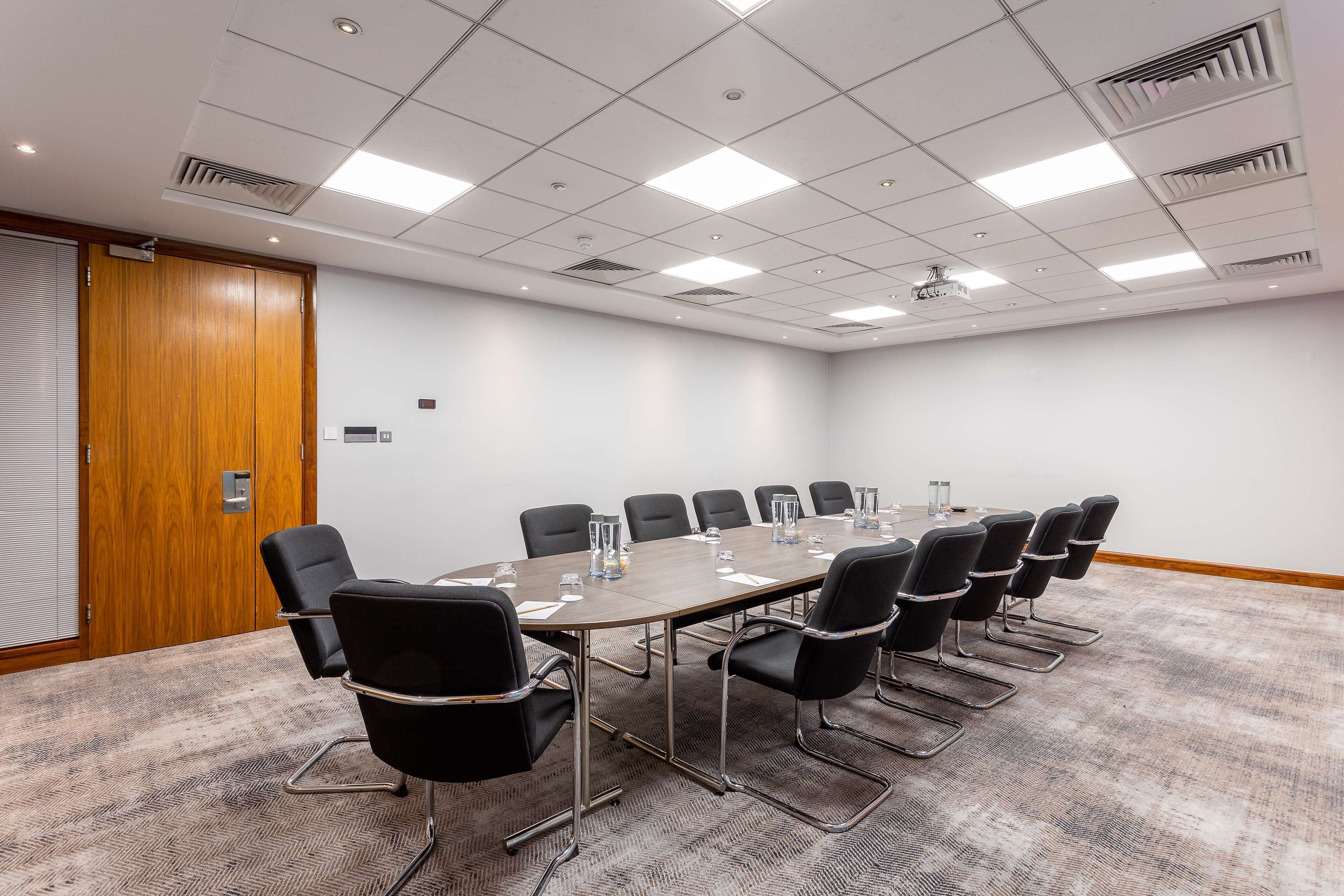 Uma sala de conferência com uma mesa comprida, cadeiras pretas, piso coberto por carpete e uma porta de madeira.