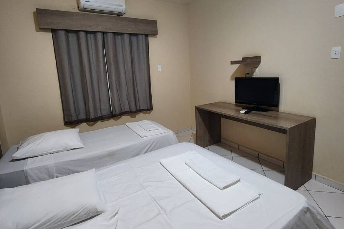Quarto de hotel com duas camas, uma escrivaninha com televisão e janela com cortinas.