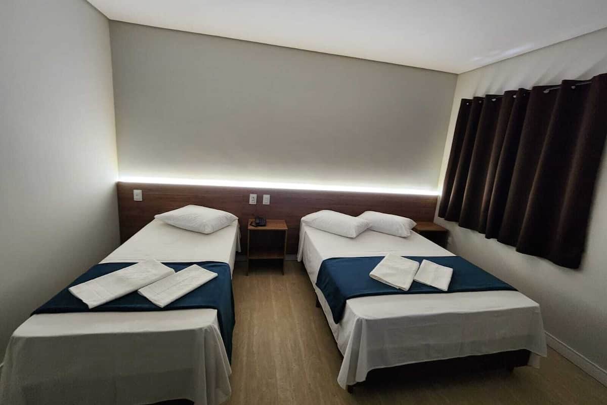 Quarto de hotel com duas camas de solteiro, mesa de cabeceira, uma pequena mesa e uma janela com cortinas.