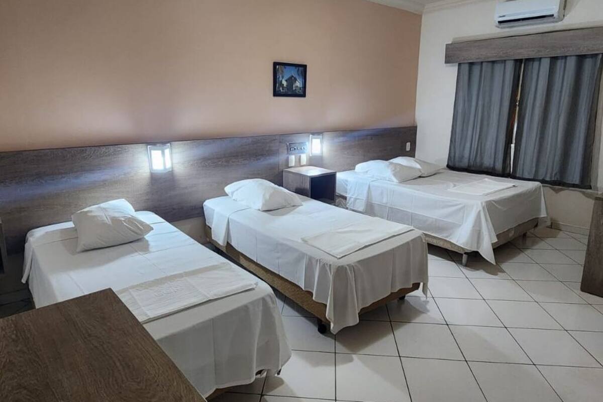 Quarto de hotel com três camas de solteiro, cabeceira de madeira, mesa de cabeceira, ar condicionado e janela com cortinas.