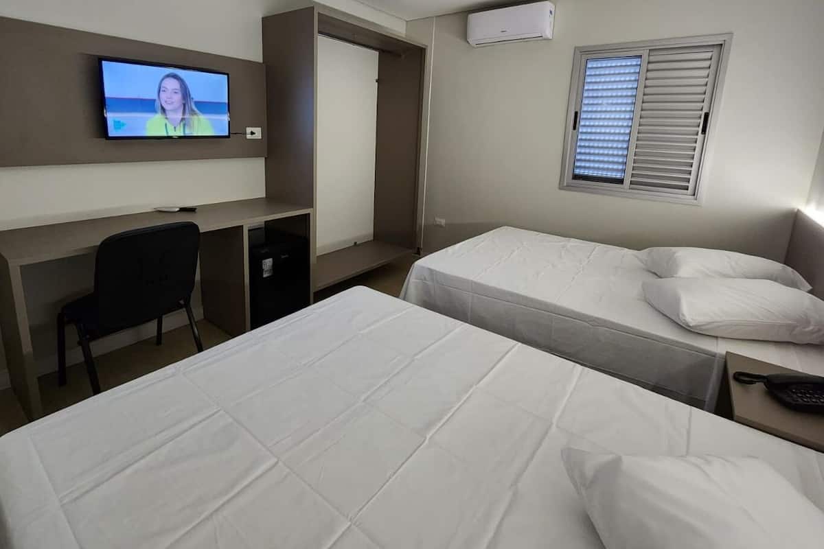 Quarto de hotel com duas camas, uma escrivaninha, uma cadeira, uma televisão e uma janela com persianas.