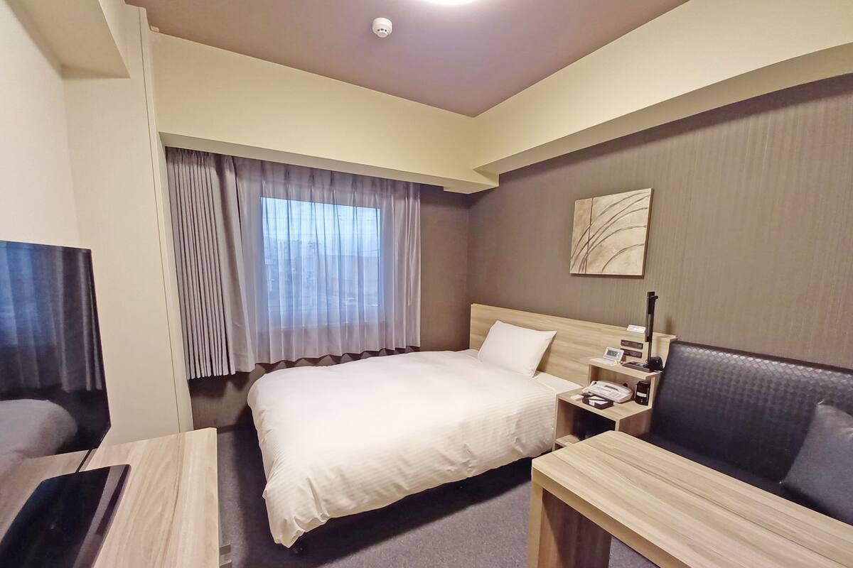 Quarto de hotel com cama, escrivaninha, cadeira, mesa de cabeceira com telefone e janela com cortinas.