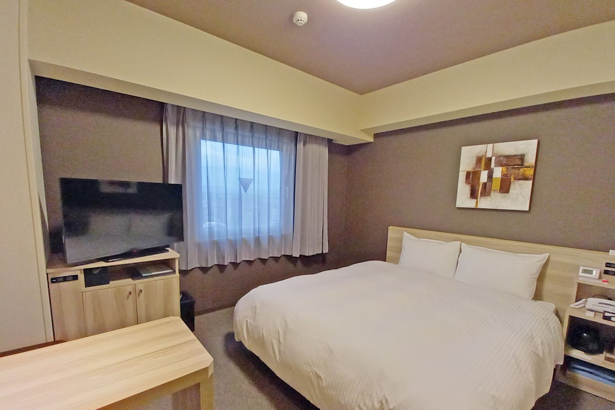 Quarto de hotel com uma cama, televisão em suporte, mesa de cabeceira, janela com cortinas e uma obra de arte na parede.