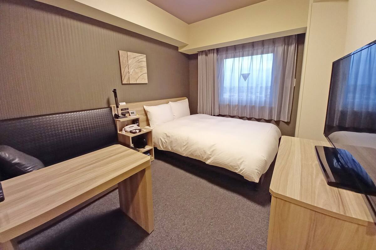 Quarto de hotel com cama, escrivaninha, cadeira, televisão e janela com cortinas.