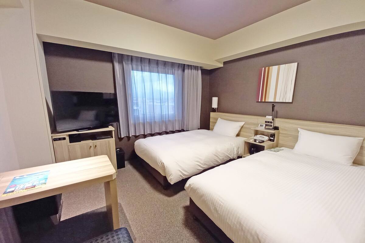 Quarto de hotel com duas camas, uma escrivaninha, televisão e janela com cortinas.