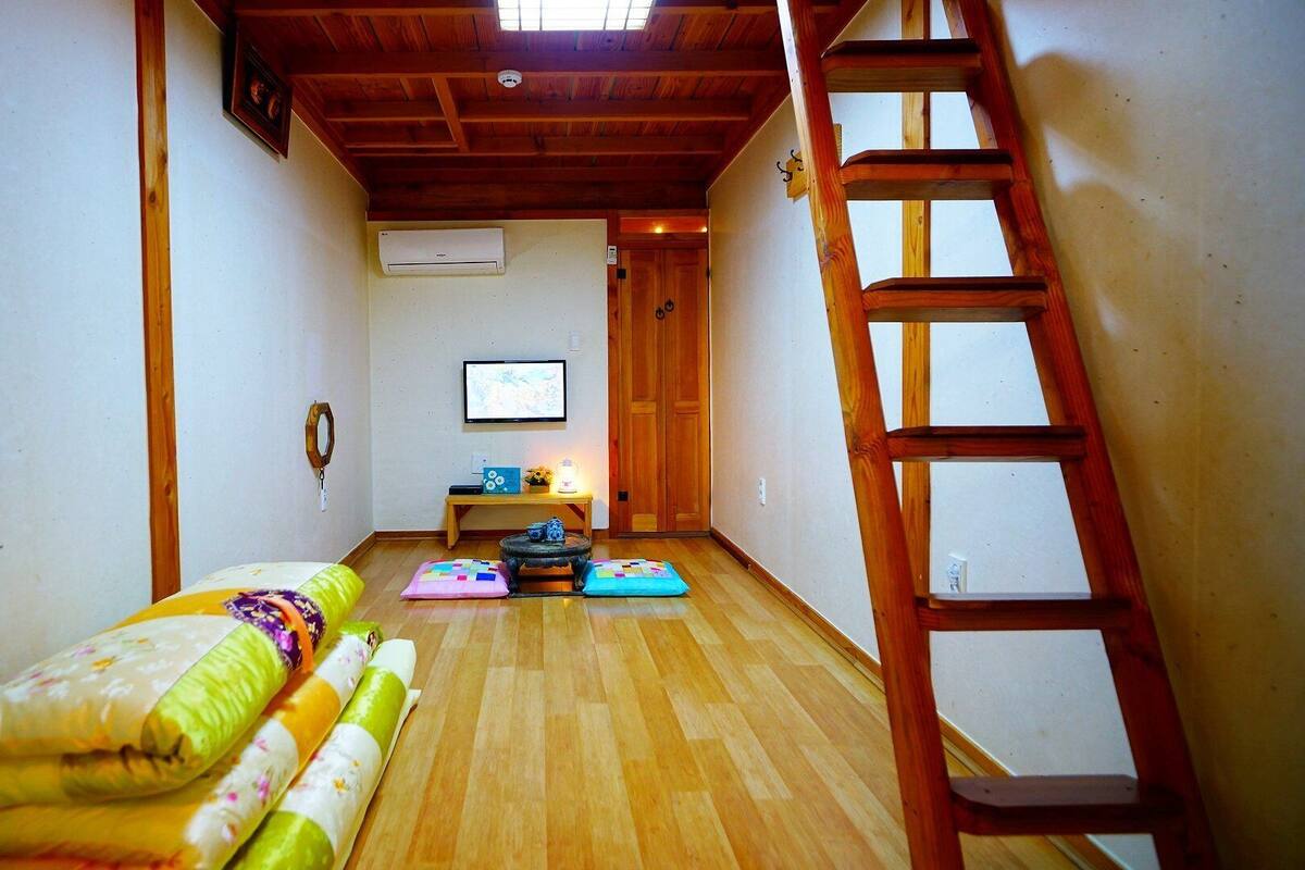 Um quarto pequeno com piso de madeira, uma cama com roupa de cama colorida, uma mesinha com uma televisão e uma escada de madeira.