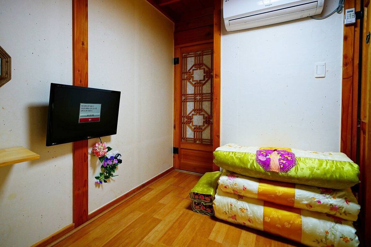 Quarto com chão de madeira, televisão fixada na parede, porta de madeira tradicional e uma pilha de roupa de cama florida.