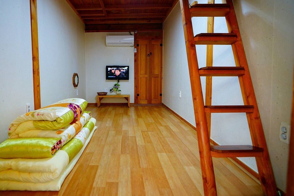 Quarto pequeno com soalho de madeira, cama com roupa de cama floral, um banco de madeira, uma televisão e uma escada de madeira.