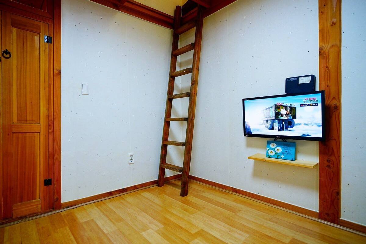 Quarto com soalho de madeira, porta de madeira, escada de madeira, televisão sobre uma prateleira de madeira e uma tomada na parede.