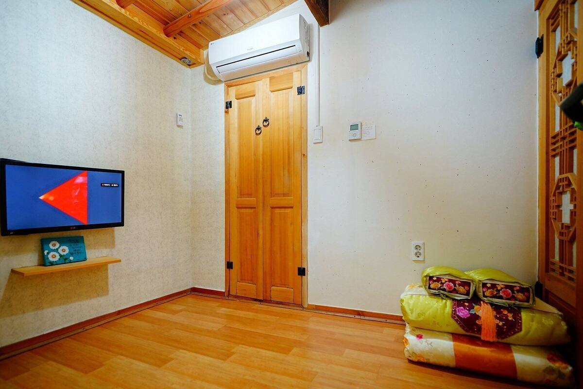 Um quarto com piso de madeira, televisão em um suporte, porta de madeira e ar-condicionado fixado na parede.