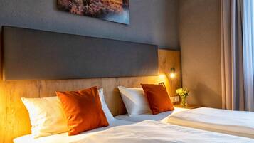 Quarto de hotel com cabeceira de madeira, duas camas com lençóis brancos, almofadas laranja e brancas, uma mesinha com um vaso de flores e um quadro na parede.