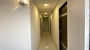 Um corredor comprido e estreito, com paredes brancas e chão de azulejos. Há luminárias no teto e um pequeno acessório decorativo na parede à esquerda. O corredor dá para uma porta com uma placa em cima.