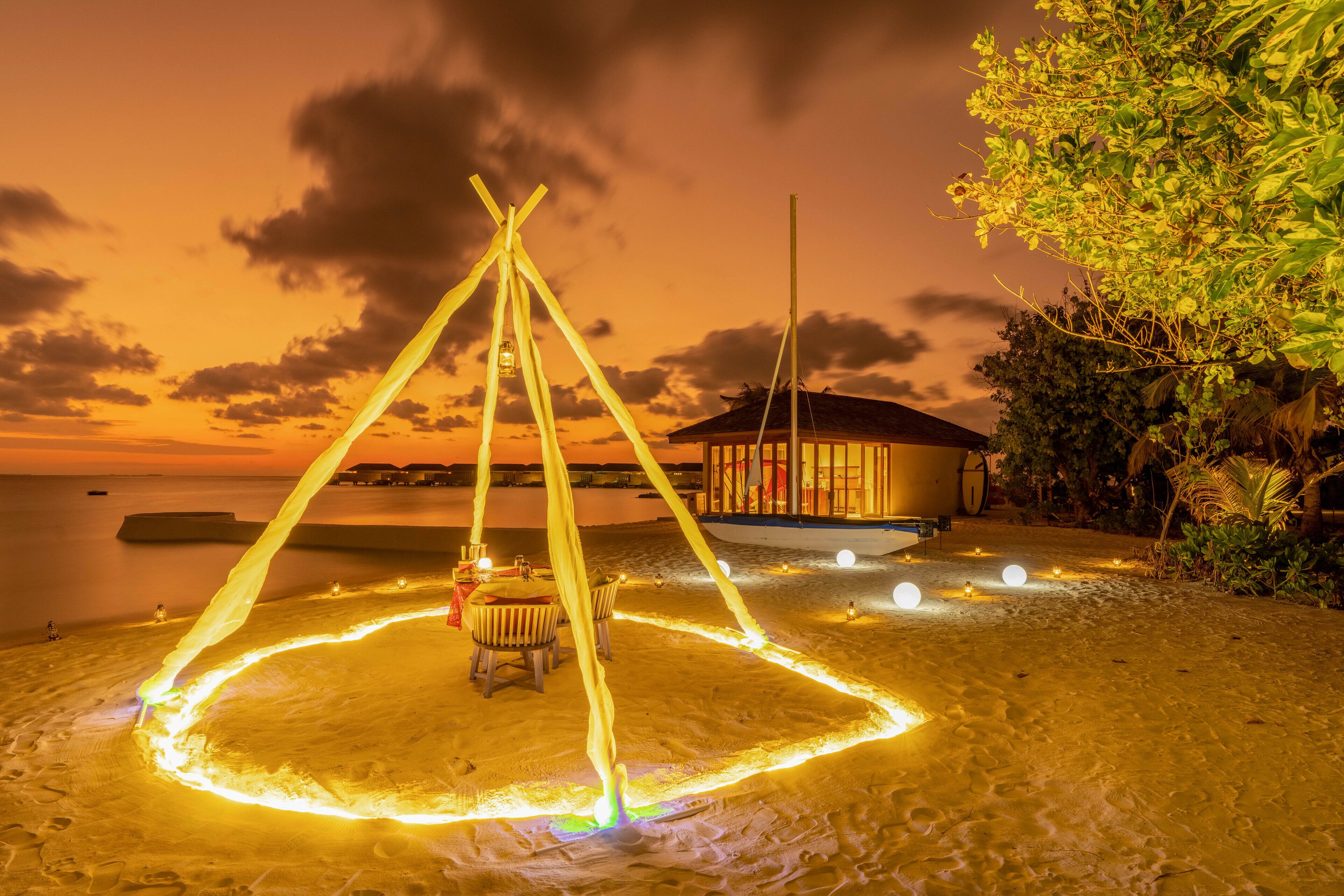 Uma praia com uma estrutura geométrica iluminada, um casebre de madeira e um barco ao fundo.