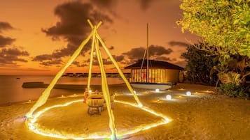 Uma praia com uma estrutura geométrica iluminada, um casebre de madeira e um barco ao fundo.