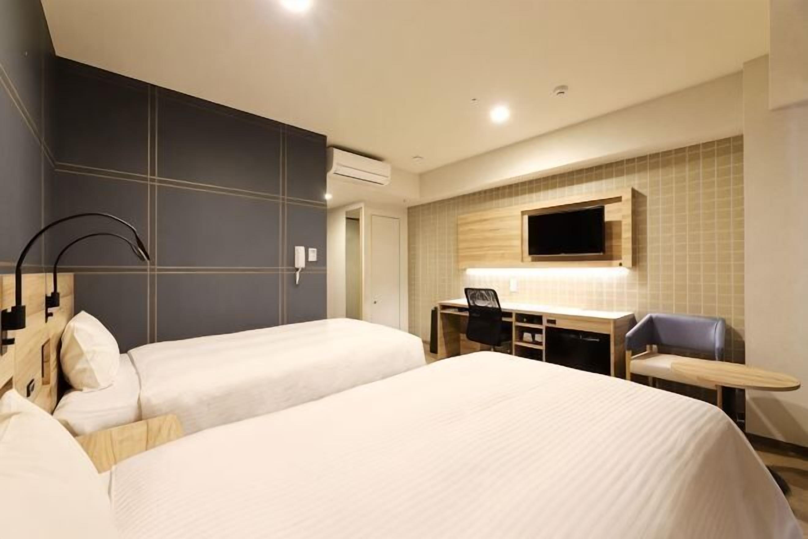 Quarto de hotel com duas camas, uma escrivaninha, uma cadeira e televisão na parede.