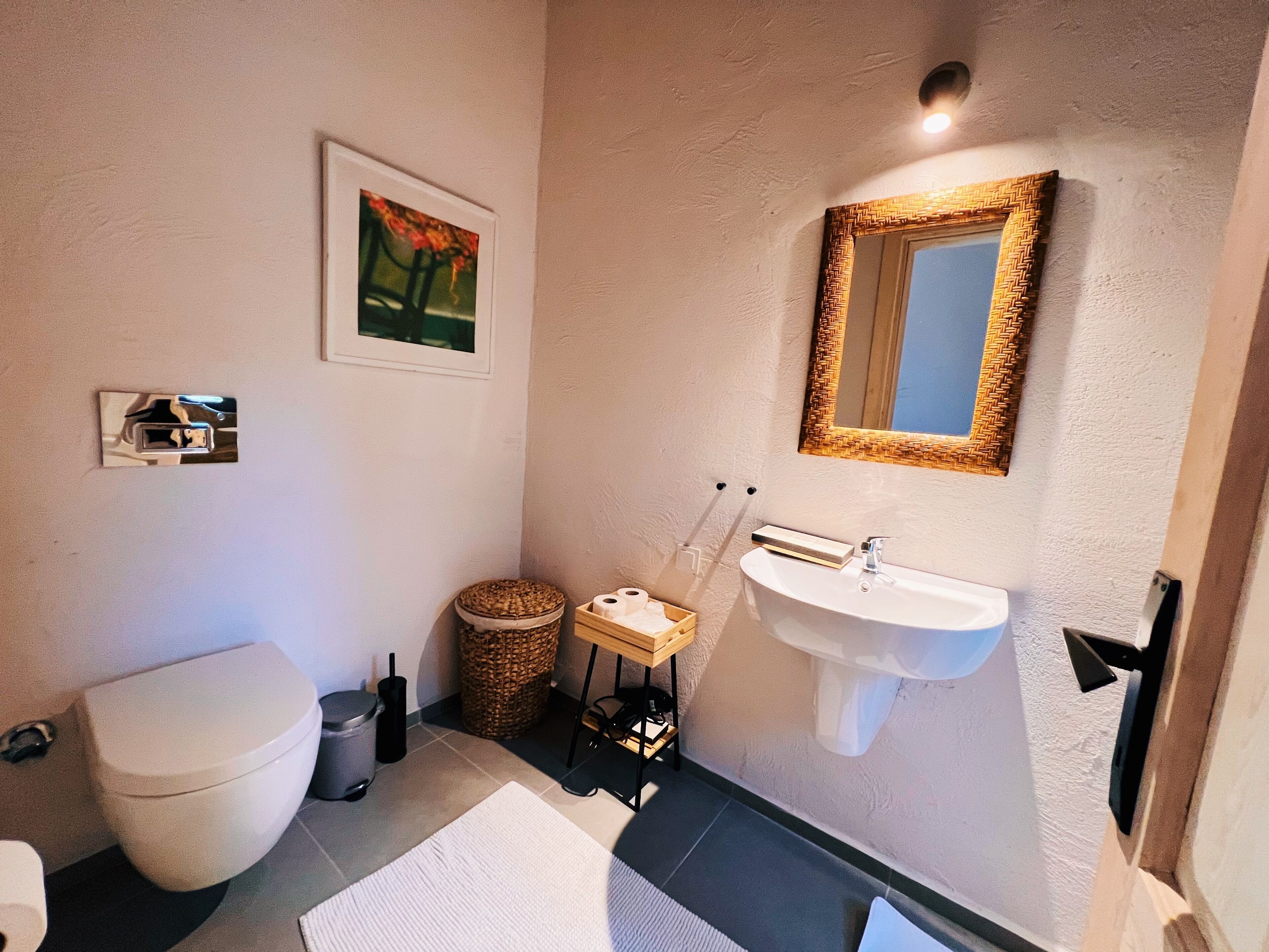 Casa de banho com sanita, lavatório, espelho, um quadro na parede e um contentor de lixo.