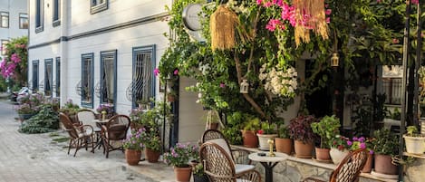 Um café à beira da rua com mesas ao ar livre, plantas em vasos e um edifício com janelas protegidas por grades.