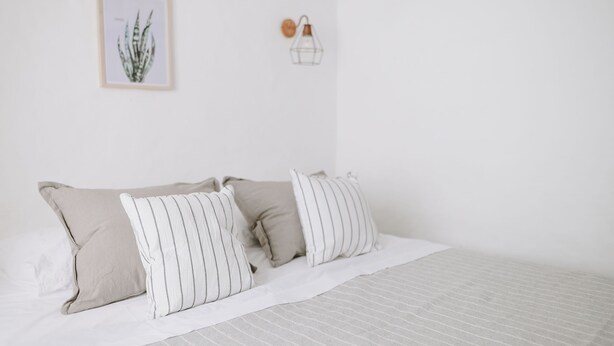 Cama bem arrumada com travesseiros listrados e uma parede branca lisa, com uma planta emoldurada e um ponto de luz fixado na parede.