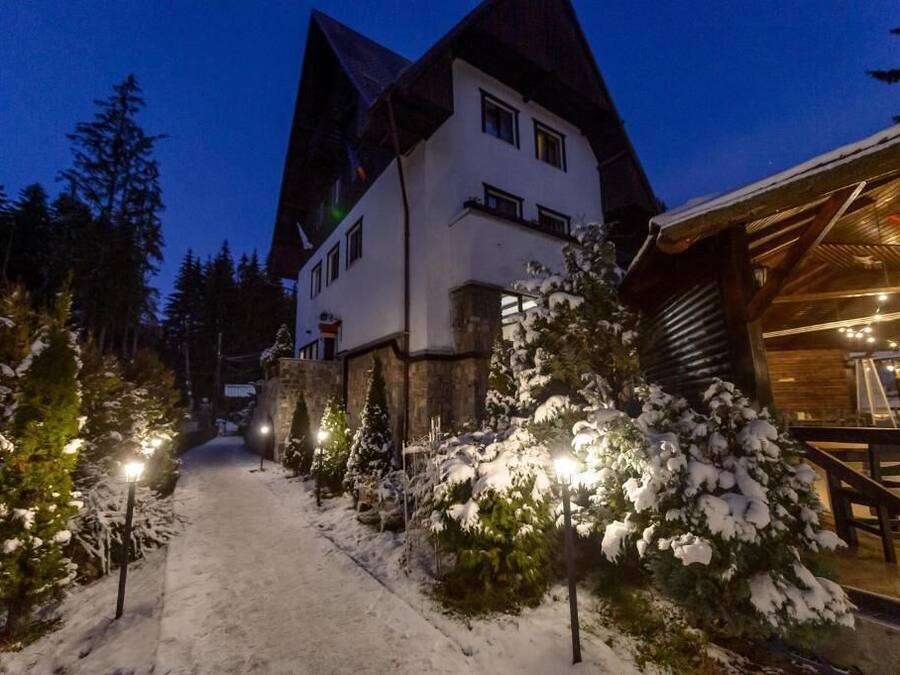 Uma trilha coberta de neve que leva a um edifício de dois andares com telhado inclinado, cercado por árvores carregadas de neve e iluminado por luzes no chão.