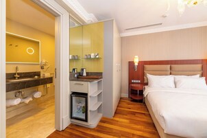 Quarto de hotel com cama, guarda-roupa embutido, mini-frigorífico, penteadeira com espelho e mesa de cabeceira com abajur.
