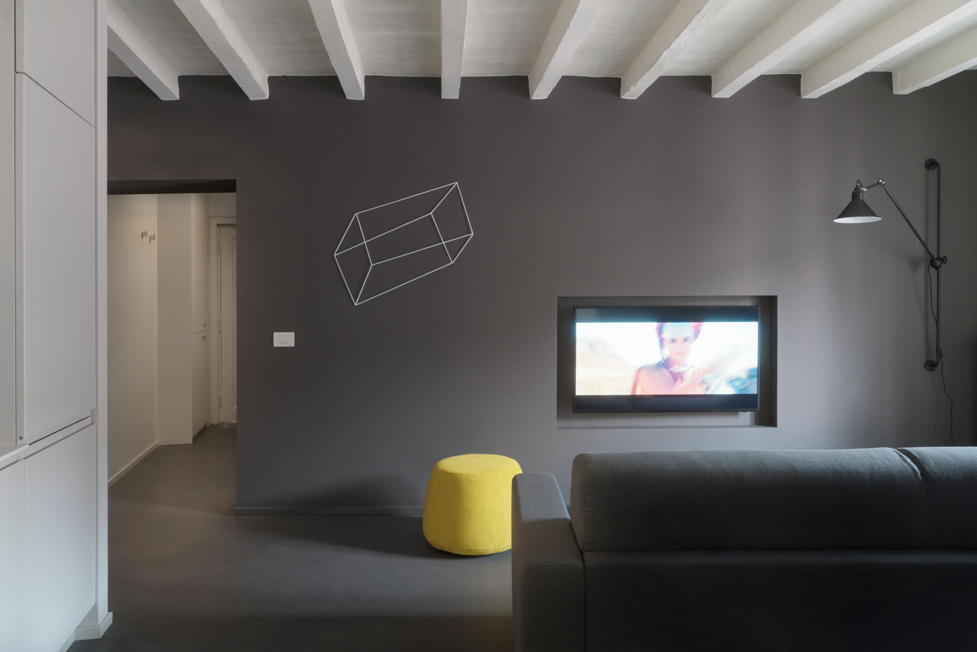 Uma sala de estar moderna com um sofá cinzento, um banquinho amarelo, uma TV de tela plana na parede e uma obra de arte geométrica.