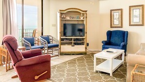 Uma sala de estar com televisão, uma poltrona azul, uma poltrona vermelha e uma varanda com cortinas.