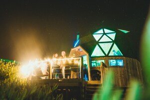 Uma cabana de madeira com telhado de vidro triangular, varanda com luzes de cordão e uma pessoa na varanda.