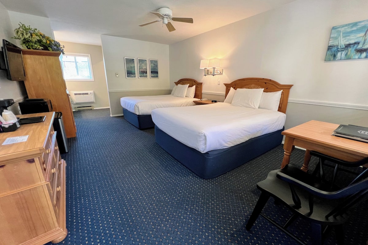 Quarto de hotel com duas camas, uma escrivaninha, uma cadeira, ventoinha no teto, abajur e um quadro na parede.