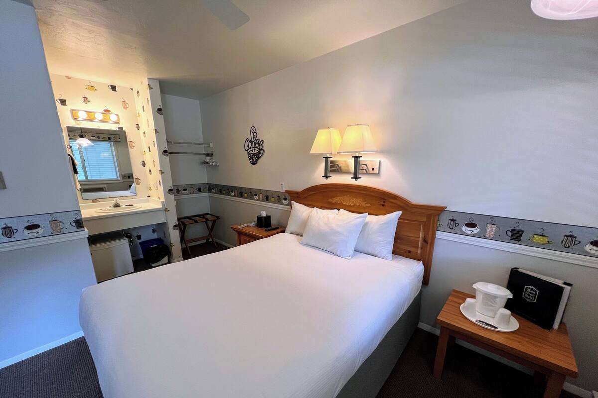 Quarto de hotel com uma cama grande, uma mesinha de cabeceira com abajur, uma pequena mesa com cafeteira e uma prateleira com objetos decorativos.