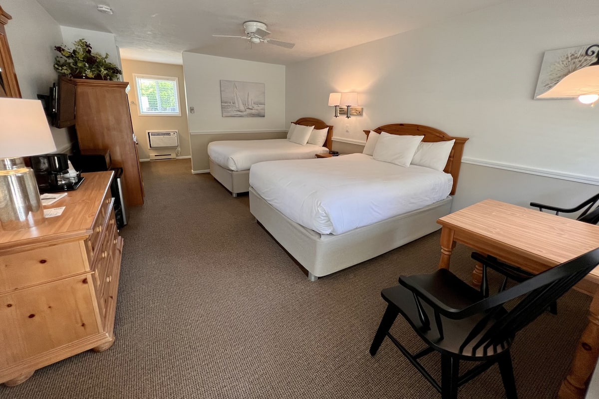 Quarto de hotel com duas camas, uma escrivaninha, uma cadeira, cafeteira, luminária, ventoinha no teto e janela com persianas.