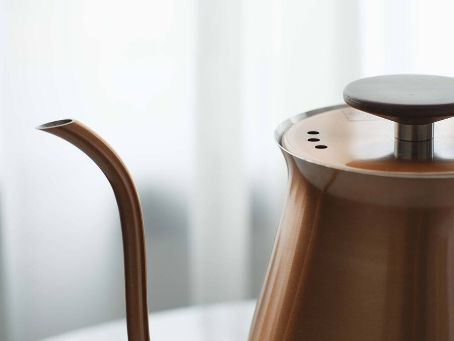 Uma cafeteira de cor cobre com bico e tampa sobre uma superfície branca.