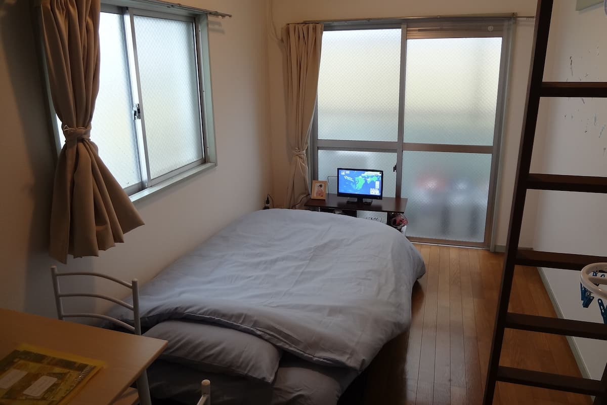 Quarto com cama, escrivaninha, cadeira, TV em móvel e janela com cortinas.