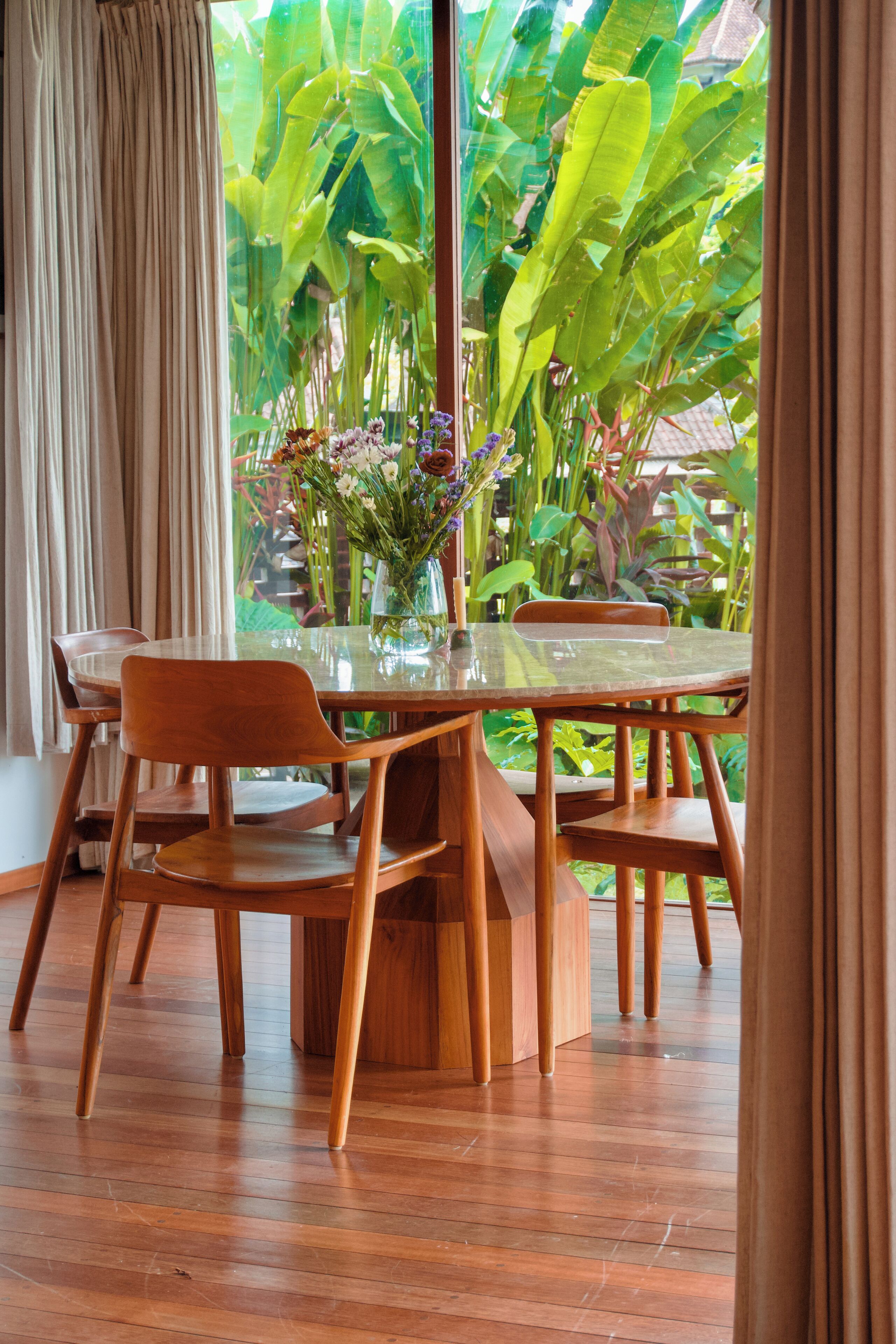 Uma área de refeições com mesa de vidro e cadeiras de madeira, um vaso com flores e vista para vegetação exuberante pela janela.