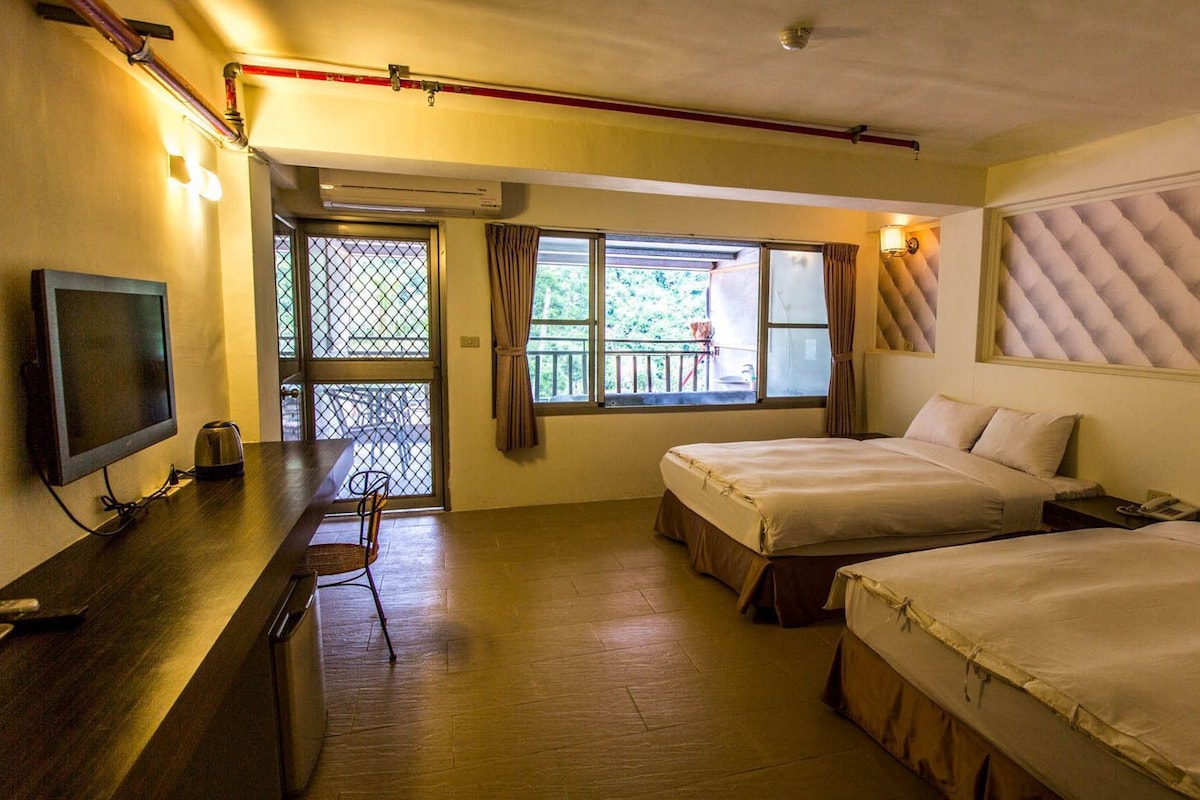 Quarto de hotel com duas camas, televisão, escrivaninha e janela com vista para o verde.