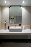 Casa de banho com lavatório branco, espelho grande e um vaso com flores.