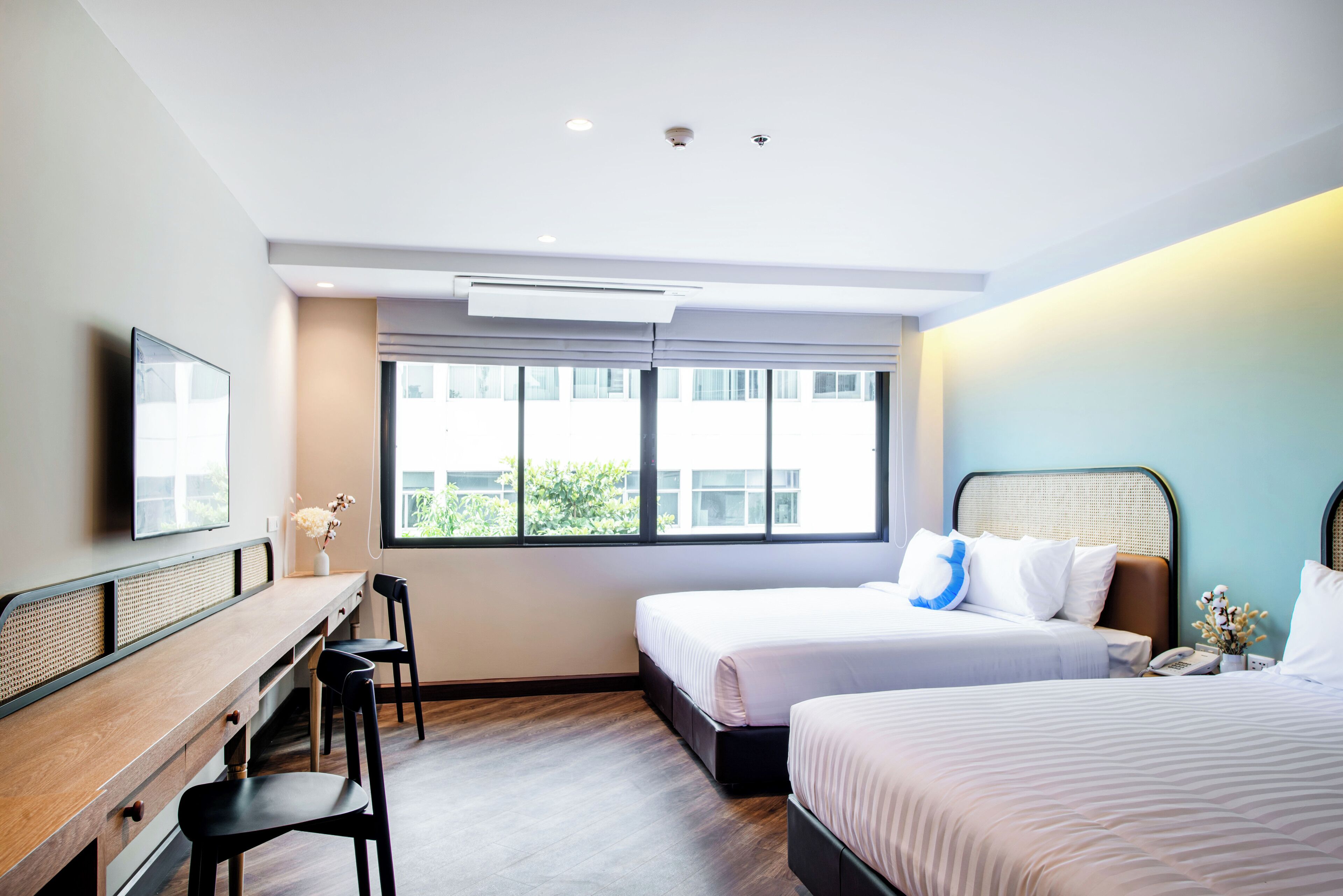 Quarto de hotel com duas camas, uma escrivaninha, cadeiras e uma janela grande.