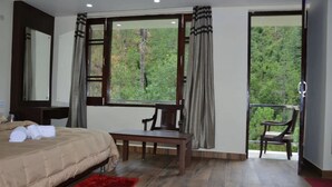Quarto com cama, escrivaninha, cadeira e vista para uma floresta através de uma janela grande.