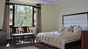 Quarto com cama grande, escrivaninha e cadeira. Há uma janela com cortinas, ventilador de teto e iluminação embutida.