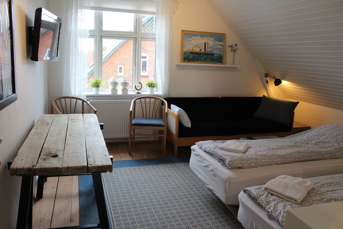 Quarto acolhedor com mesa de madeira, duas camas de solteiro, um sofá e uma TV fixada na parede.