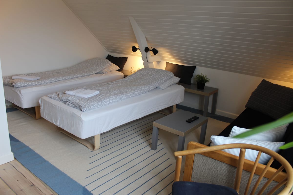 Um quarto pequeno e acolhedor com duas camas de solteiro, uma mesinha, uma cadeira e um abajur.