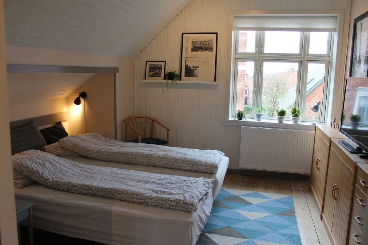 Quarto com duas camas, janela com vista para edifícios, radiador e escrivaninha com luminária.