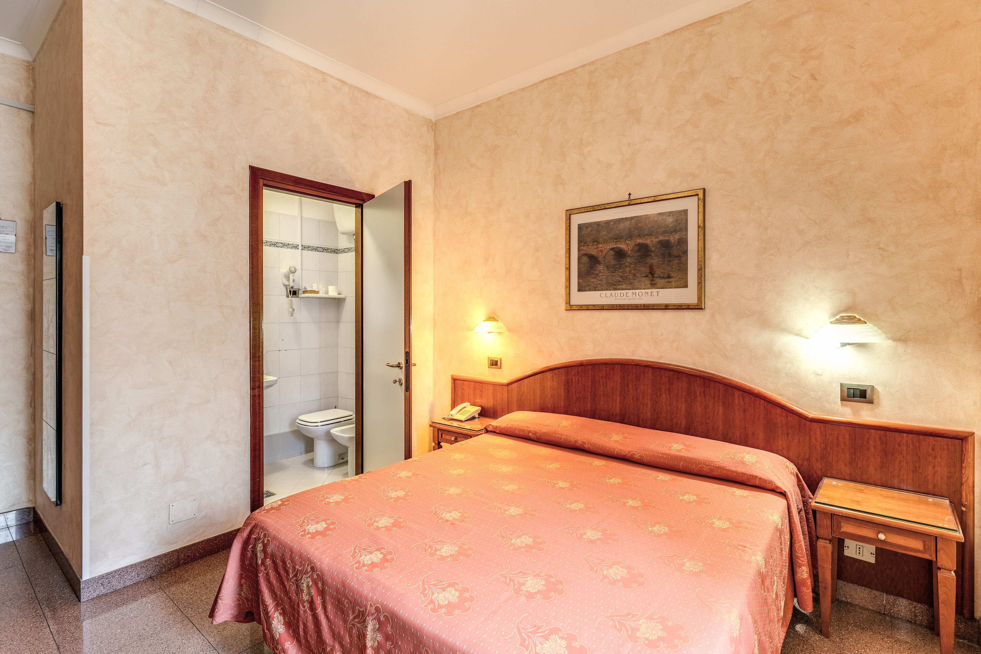 Quarto de hotel com cama, mesa de cabeceira, luminária na parede, quadro emoldurado e banheiro visível por uma porta aberta.
