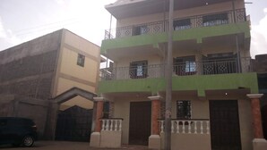Um edifício residencial de vários andares com fachada verde e bege, duas varandas visíveis e entrada com portão.