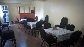 Uma sala de jantar com várias mesas e cadeiras, uma área de cozinha ao fundo e uma janela com cortinas.