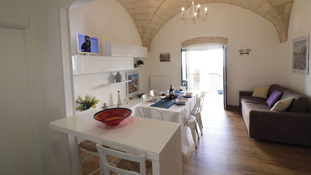 Ampla sala de estar com uma mesa de jantar posta para quatro pessoas, um sofá e um lustre.
