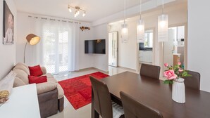 Uma sala de estar moderna com área de jantar, televisão de tela plana e cozinha visível através de uma porta aberta.
