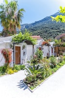Um edifício branco com porta de madeira, rodeado por vegetação exuberante e uma montanha ao fundo.