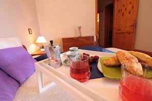 Cama bem arrumada com travesseiro roxo, mesa de cabeceira com abajur, bandeja com comida e bebidas, e porta de madeira ao fundo.