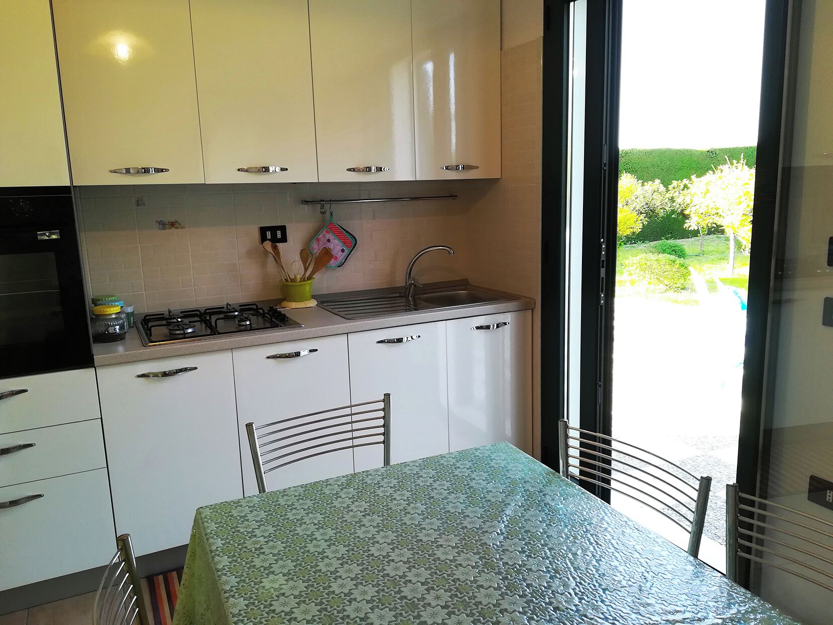 Cozinha compacta com fogão a gás, pia e armários brancos. Há uma mesa de jantar com toalha de mesa estampada e uma cadeira.
